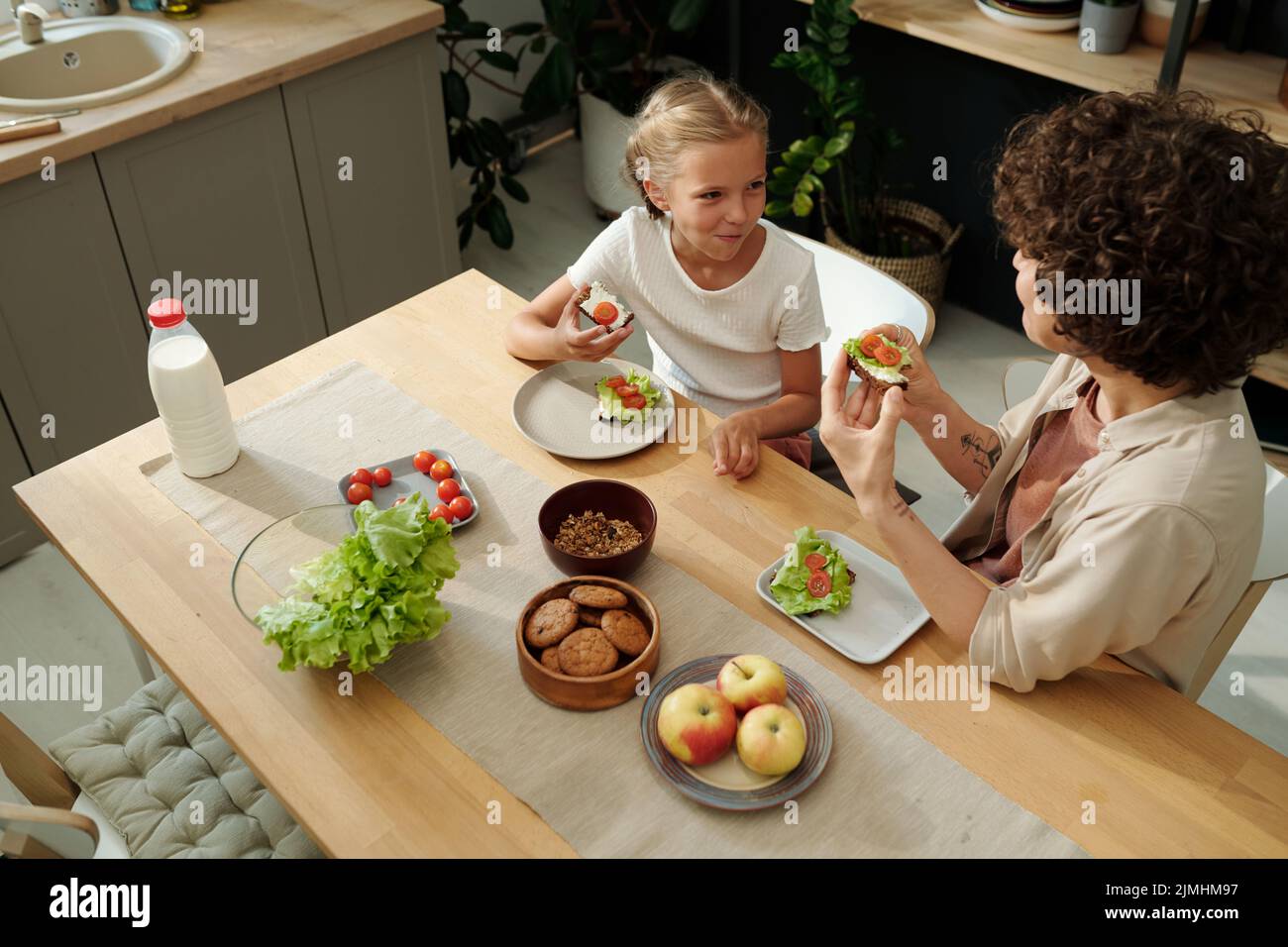 Happy youthful girl and her mother eating fresh vegetable sandwiches for breakfast while sitting ...