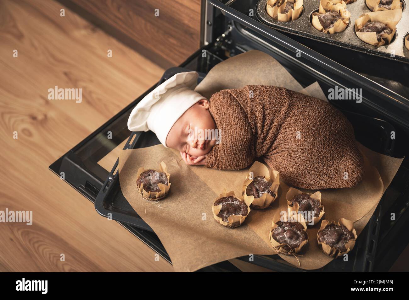Newborn baby wearing chef's hat is lying on the oven tray with a ...