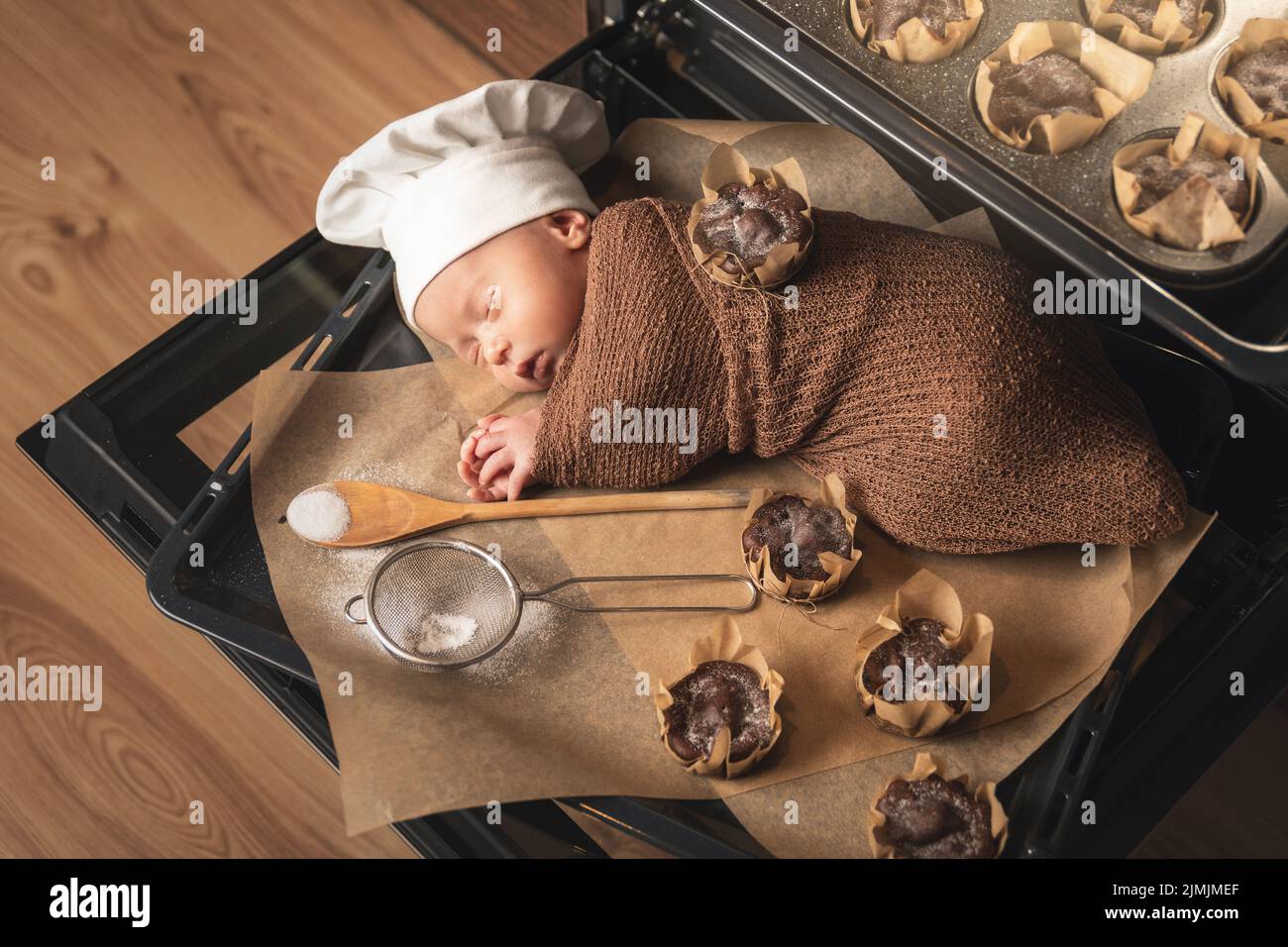 Newborn baby wearing chef's hat is lying on the oven tray with a ...