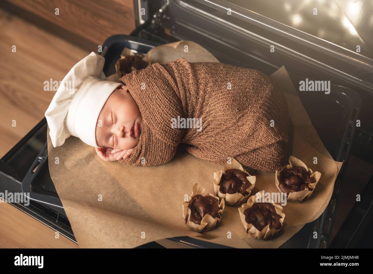 Newborn baby wearing chef's hat is lying on the oven tray with a ...