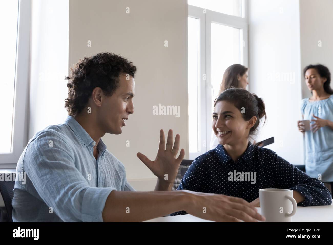 Hispanic and Indian colleagues talking seated at desk at workplace ...