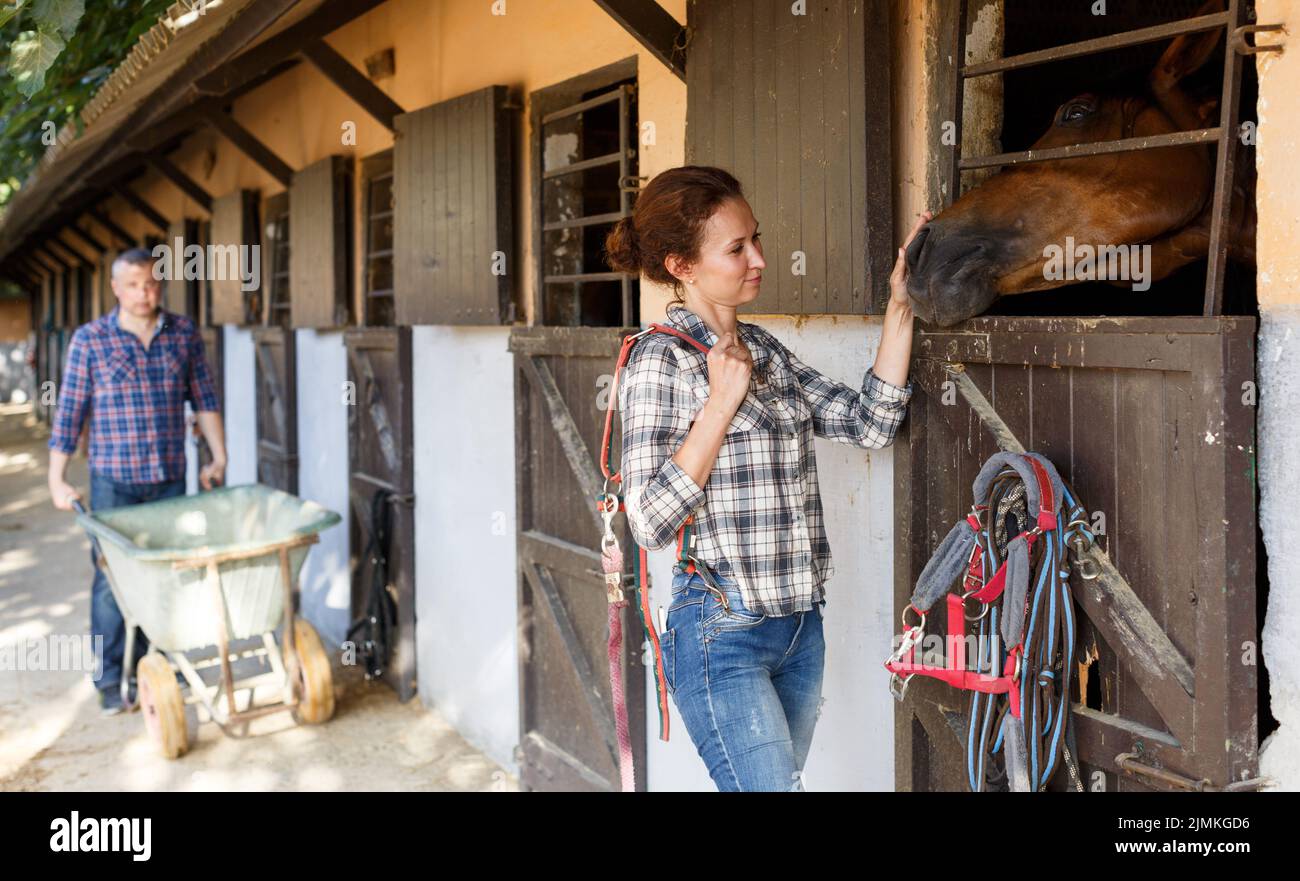 Mature positive female farmer standing near horse at stable Stock Photo ...