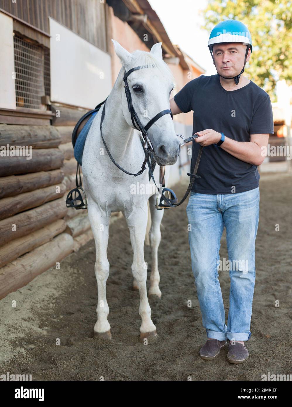 Mature smiling man farmer in helmet standing with white horse at stable ...