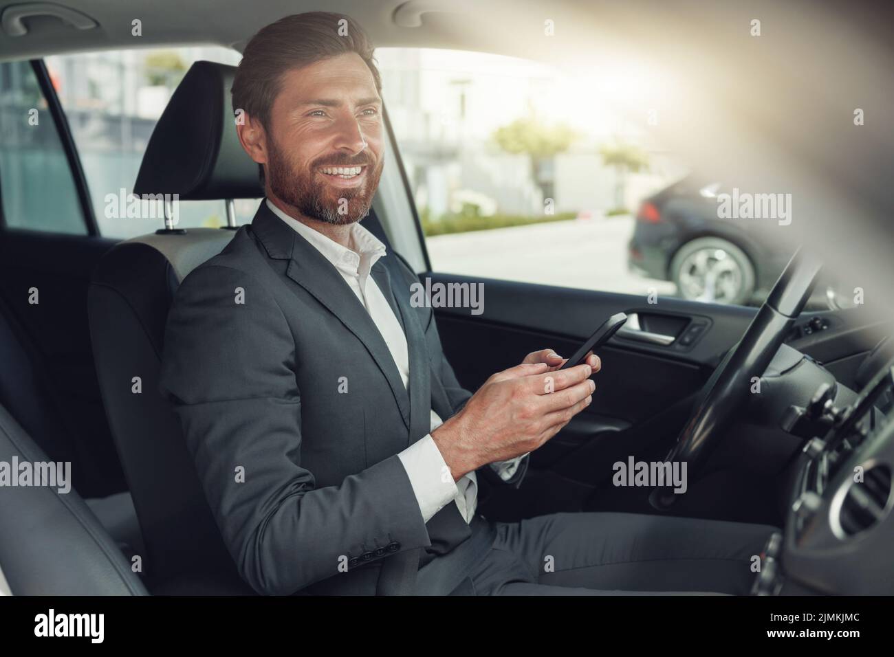 Smiling businessman in grey suit is riding behind steering wheel of car ...
