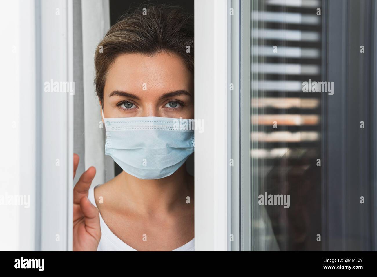 Scared woman wearing prevention mask looking outside through the window ...