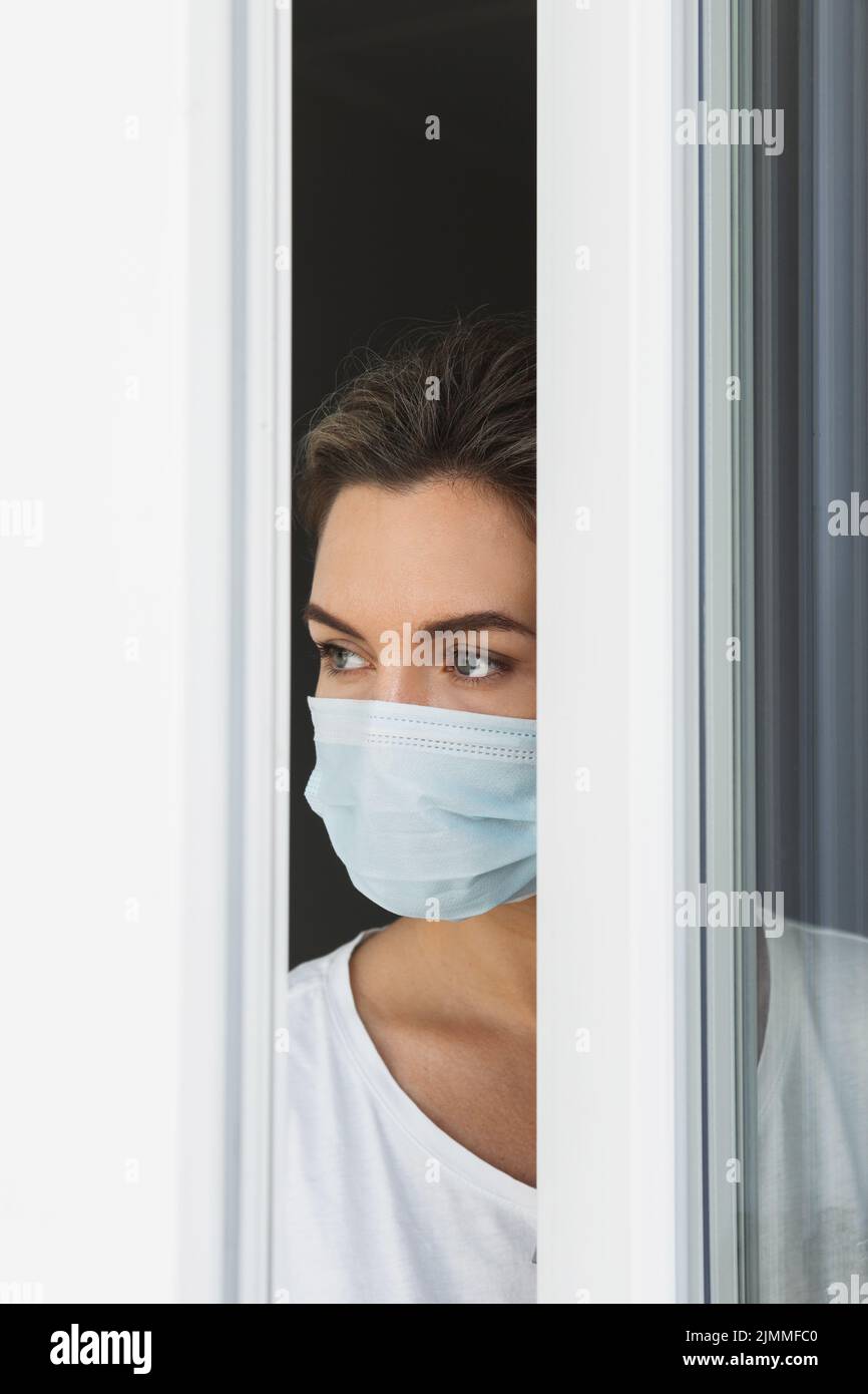 Scared woman wearing prevention mask looking outside through the window ...