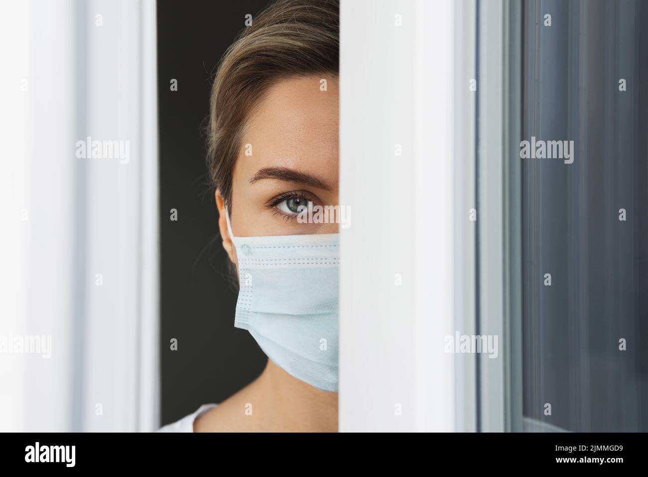 Scared woman wearing prevention mask looking outside through the window ...