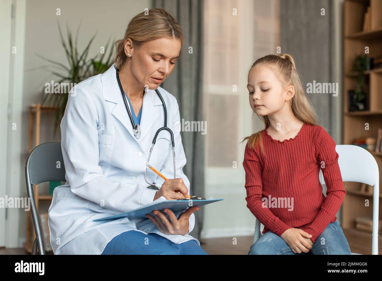 Focused female doctor writing down girl's symptoms on clipboard while ...