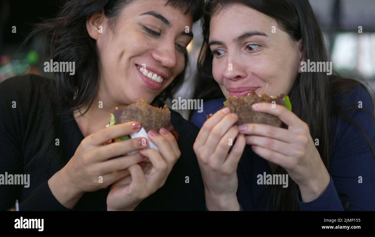 Two happy female friends eating burgers at the same time. Young women ...