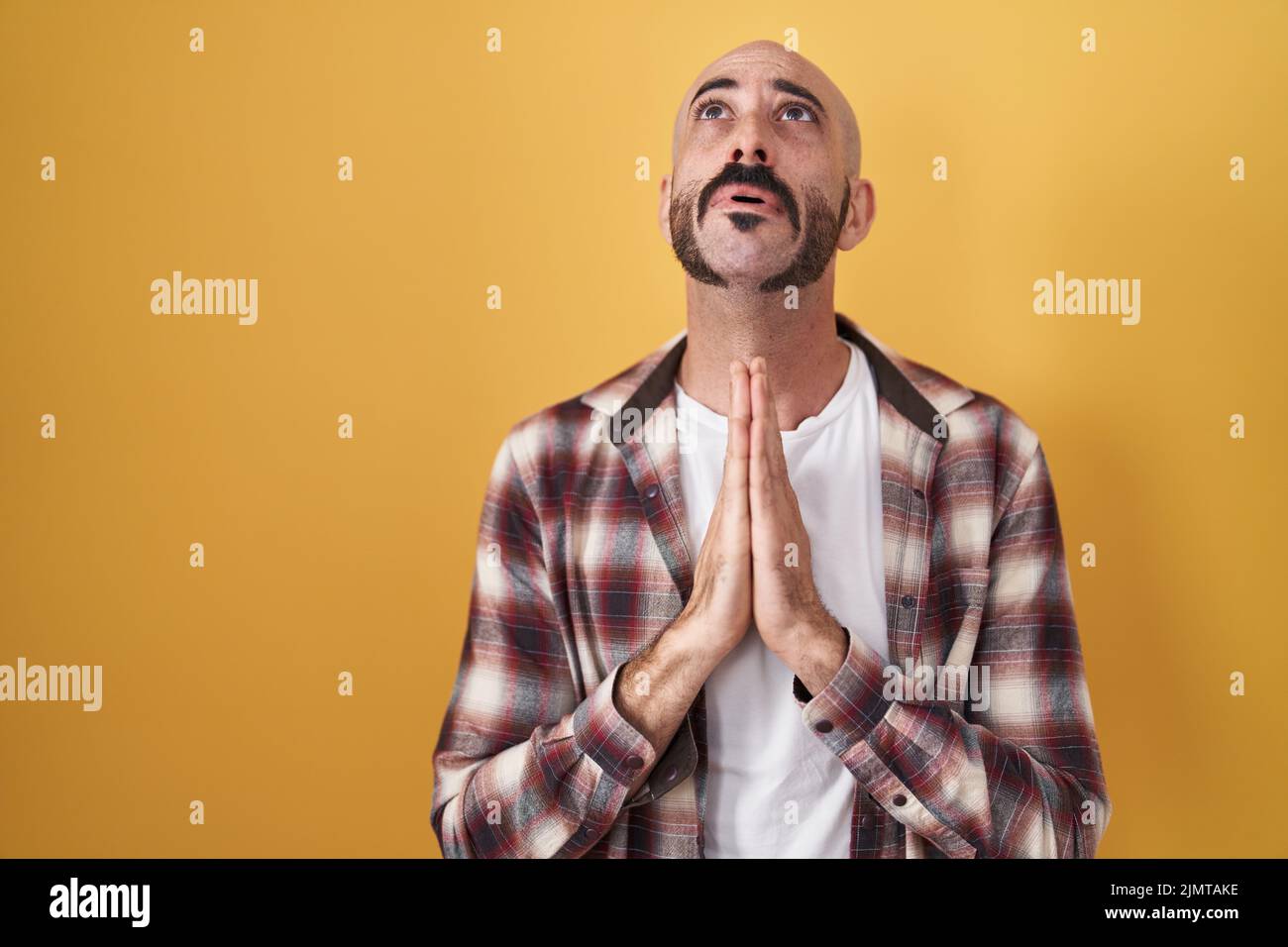 Hispanic man with beard standing over yellow background begging and ...