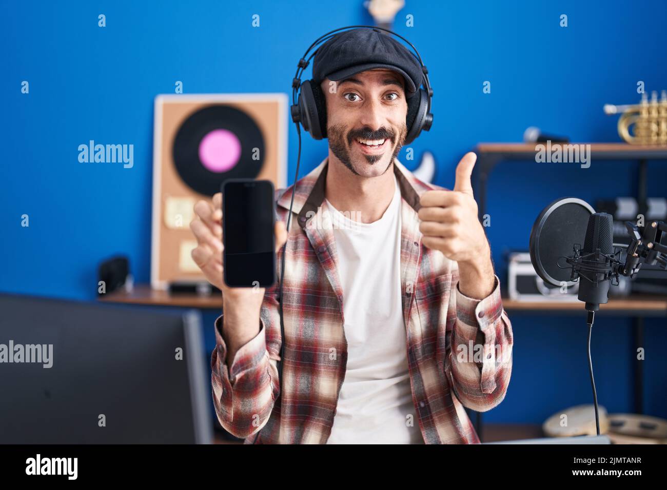Hispanic man with beard at music studio showing smartphone smiling ...