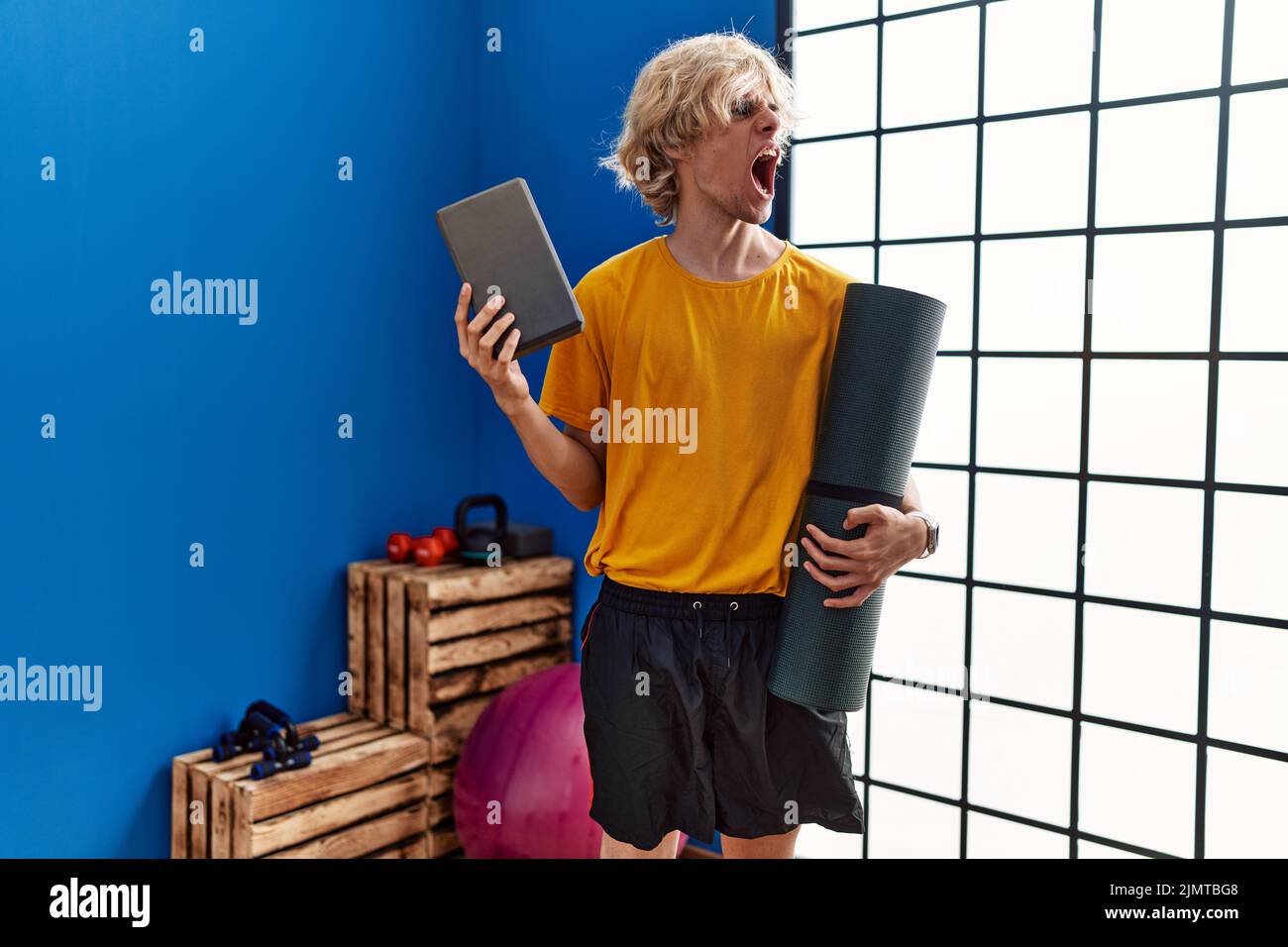 Young man holding yoga room angry and mad screaming frustrated and ...