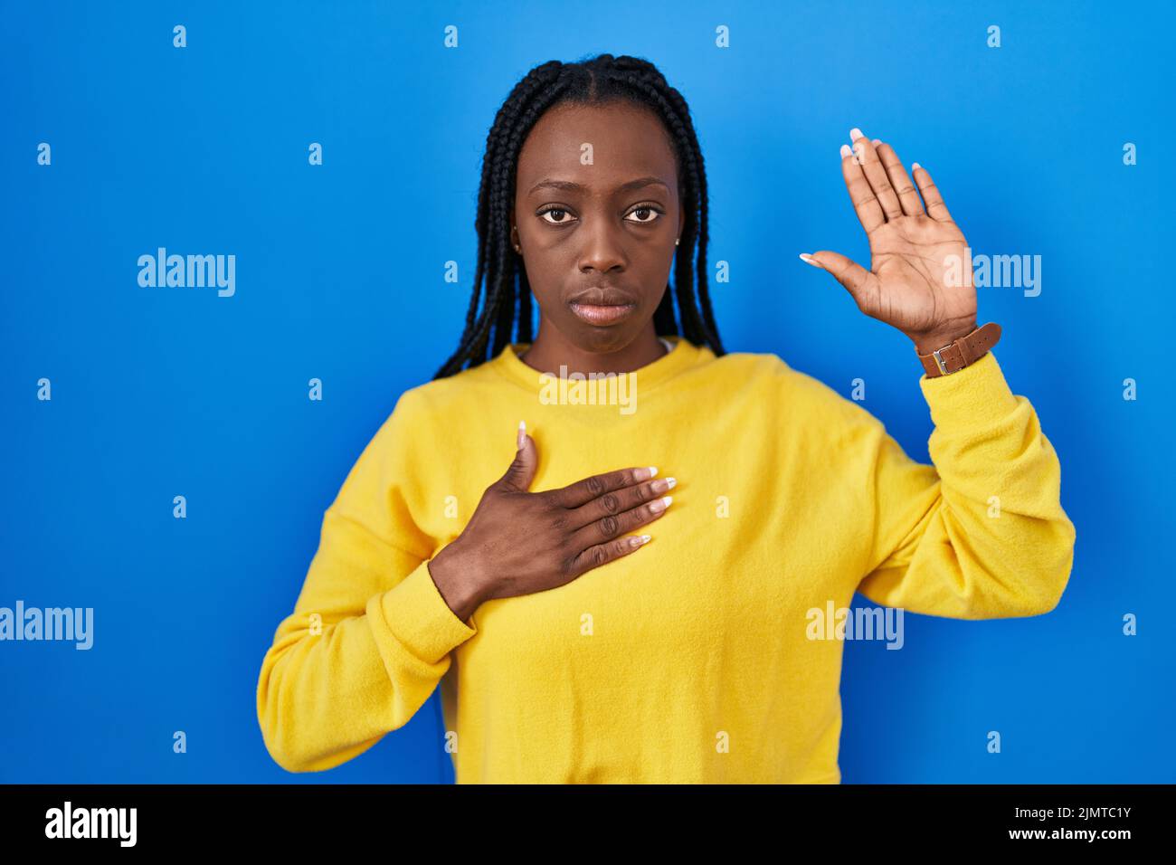 Beautiful black woman standing over blue background swearing with hand ...