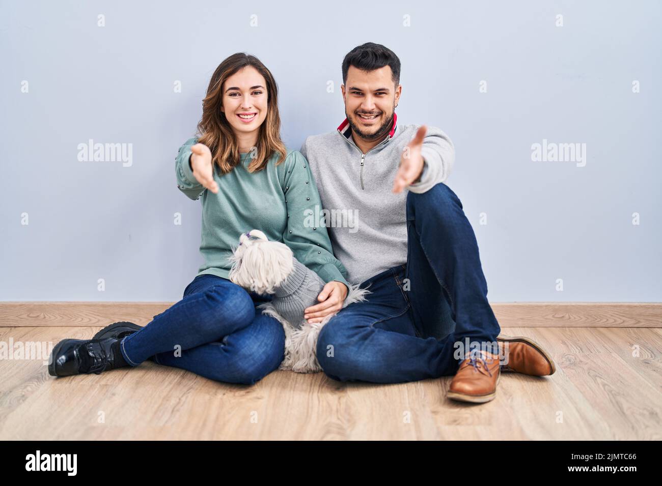 Young hispanic couple sitting on the floor with dog smiling friendly offering handshake as ...