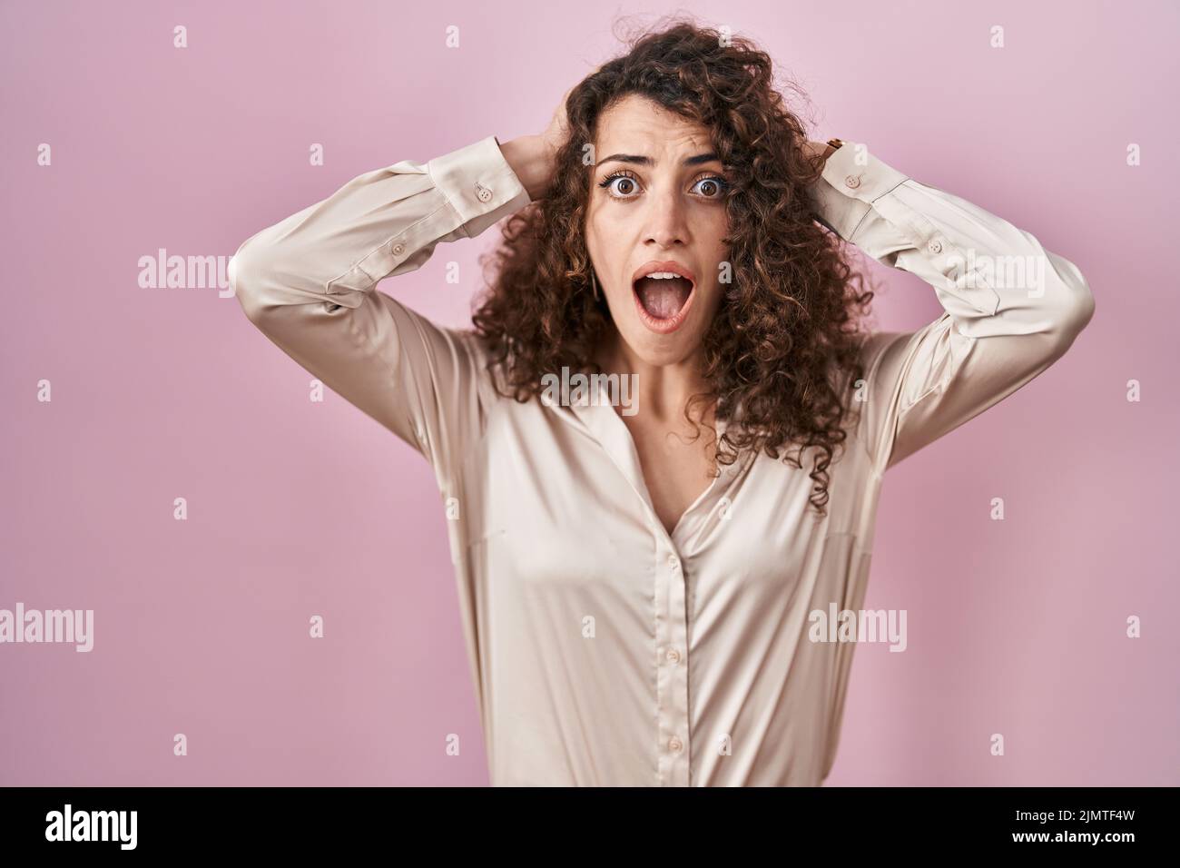 Hispanic woman with curly hair standing over pink background crazy and ...