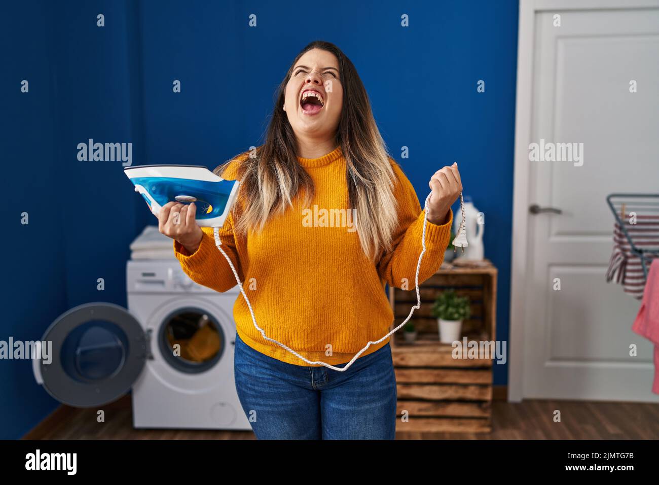 Young hispanic woman holding electric iron angry and mad screaming ...