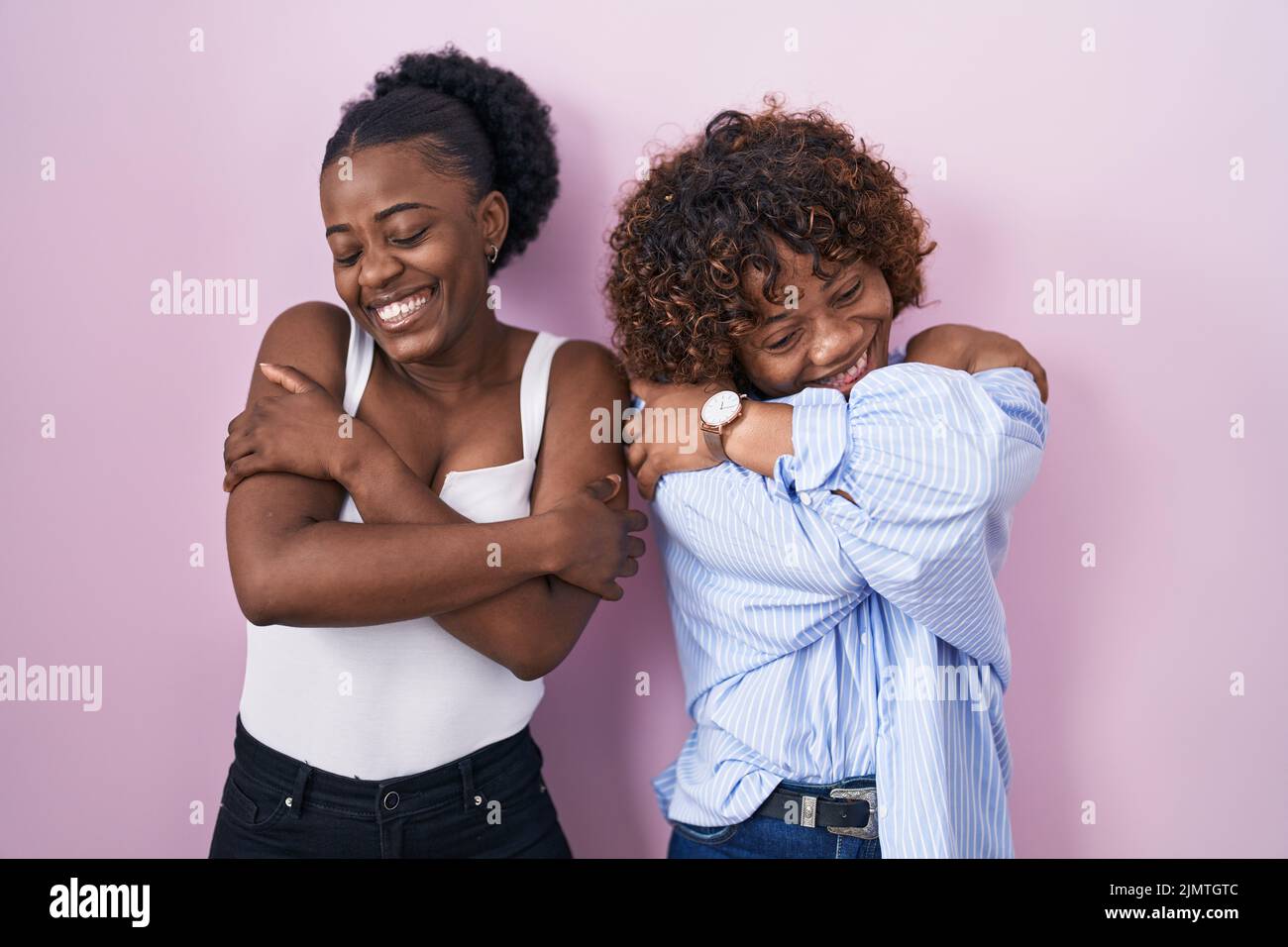 Two african women standing over pink background hugging oneself happy ...
