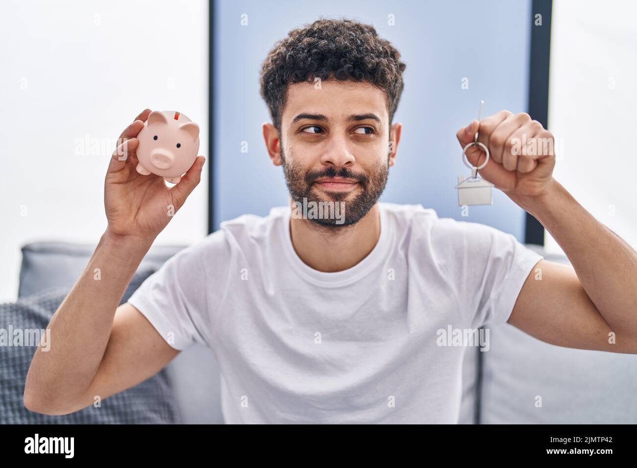 Arab man holding piggy bank and house keys smiling looking to the side ...