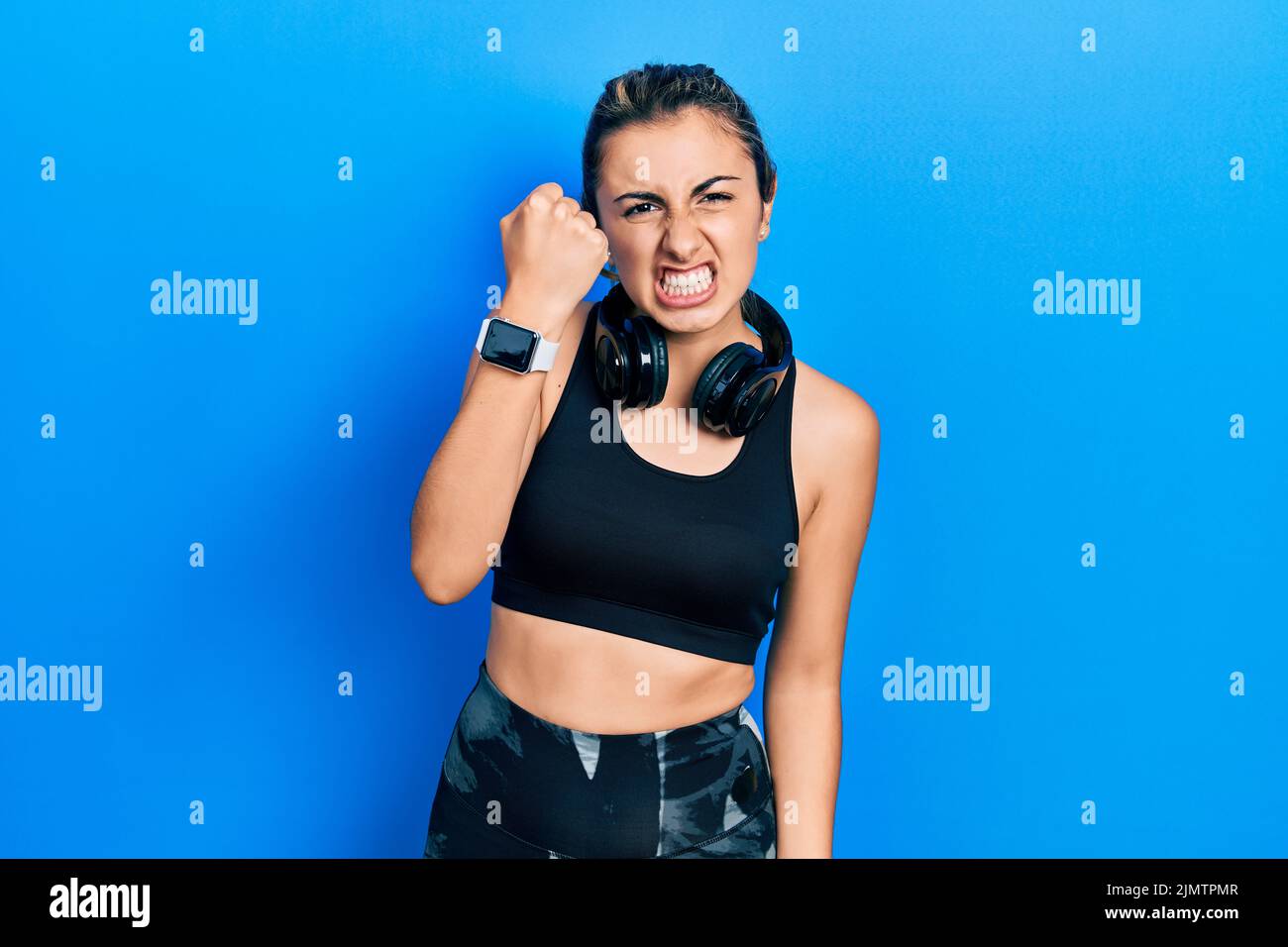 Beautiful hispanic woman wearing gym clothes and using headphones angry ...