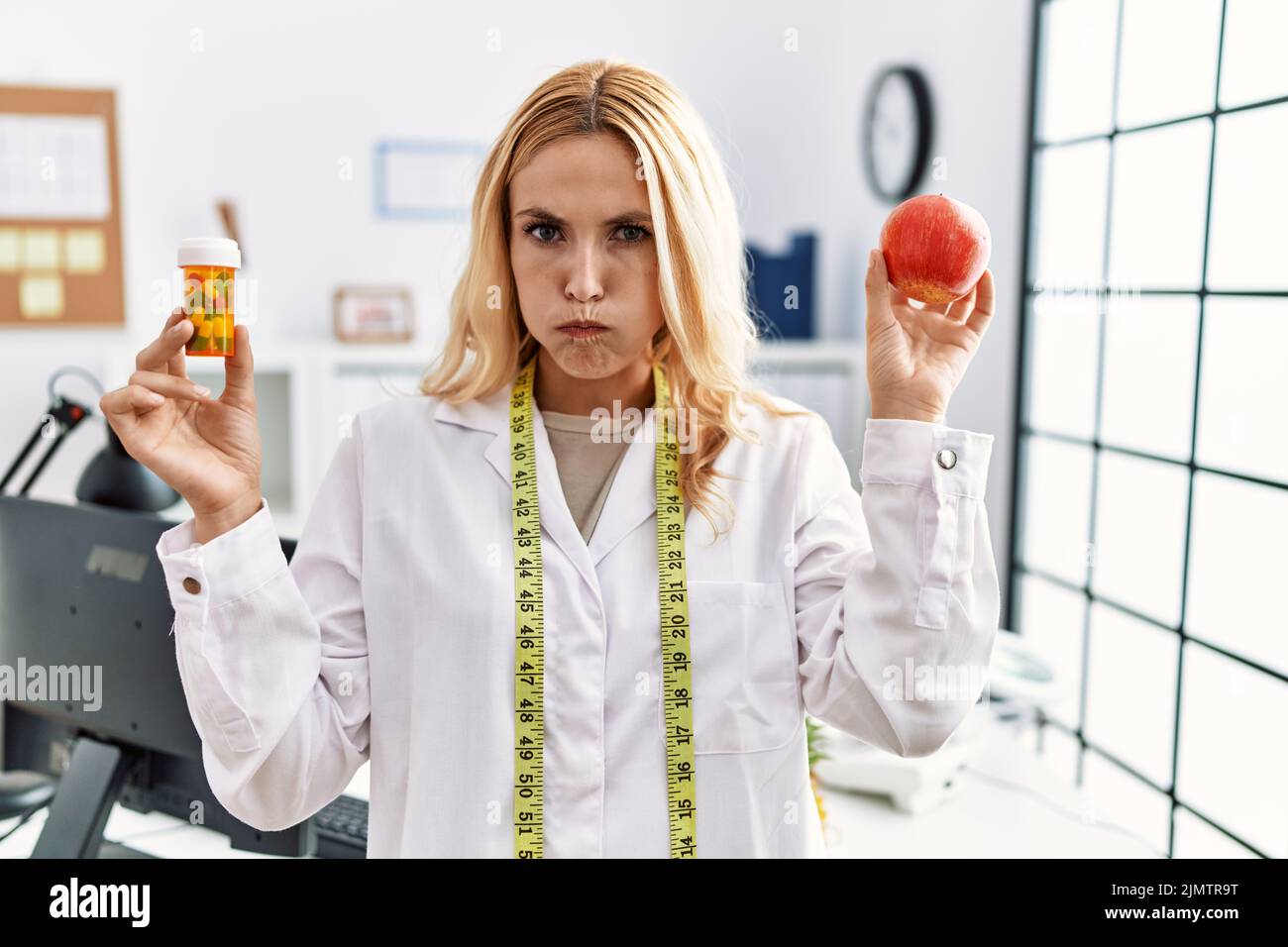 Beautiful blonde nutritionist woman holding fat pills puffing cheeks ...