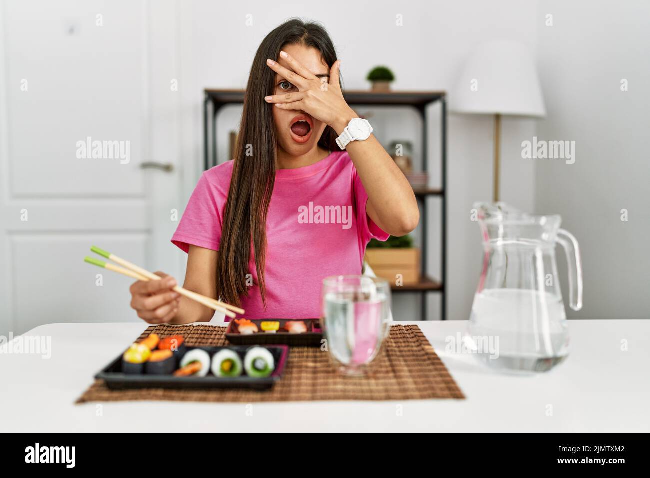 Young brunette woman eating sushi using chopsticks peeking in shock ...
