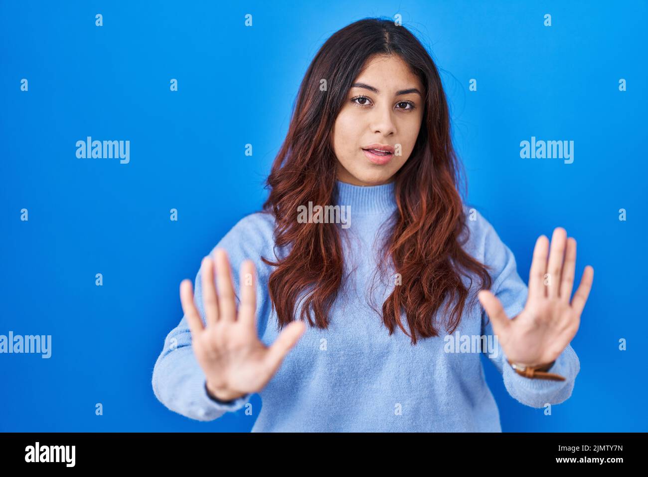 Hispanic young woman standing over blue background moving away hands ...