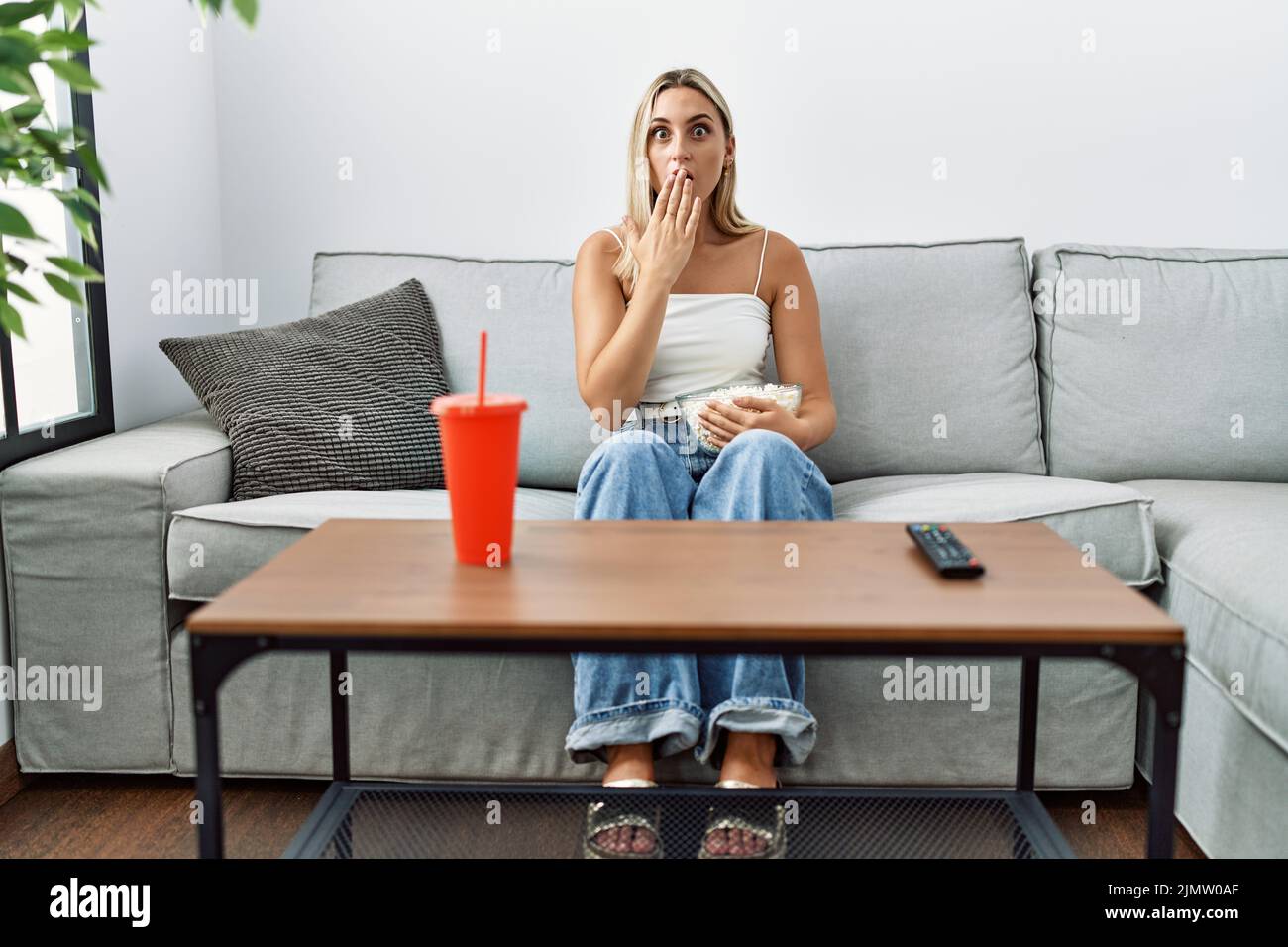 Young blonde woman eating popcorn sitting on the sofa covering mouth ...