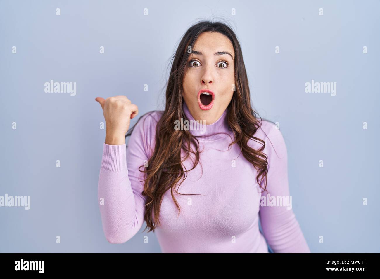 Young brunette woman standing over blue background surprised pointing ...