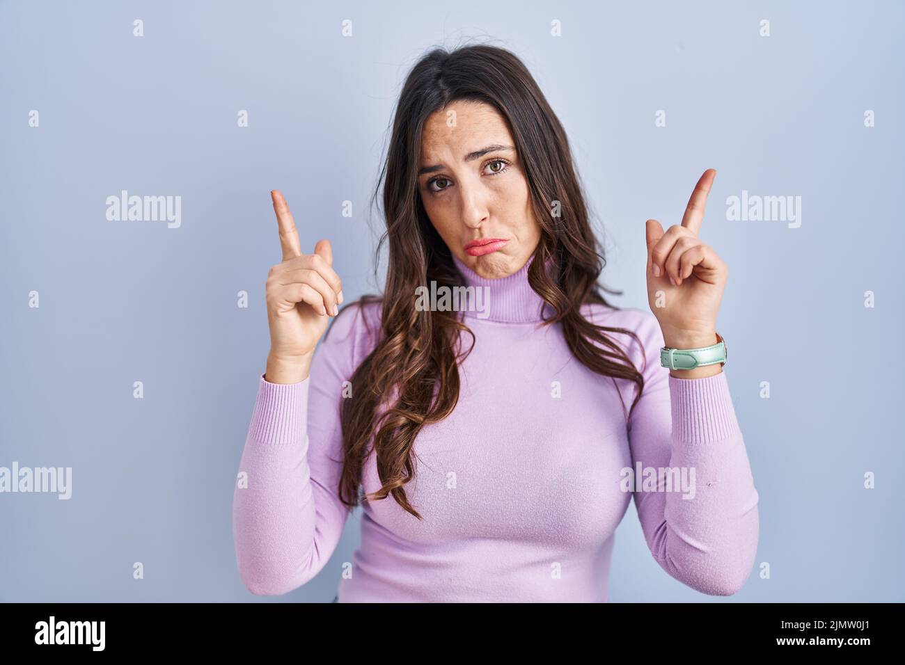 Young brunette woman standing over blue background pointing up looking ...