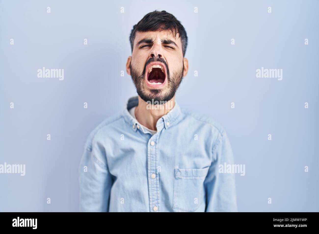 Young hispanic man with beard standing over blue background angry and ...