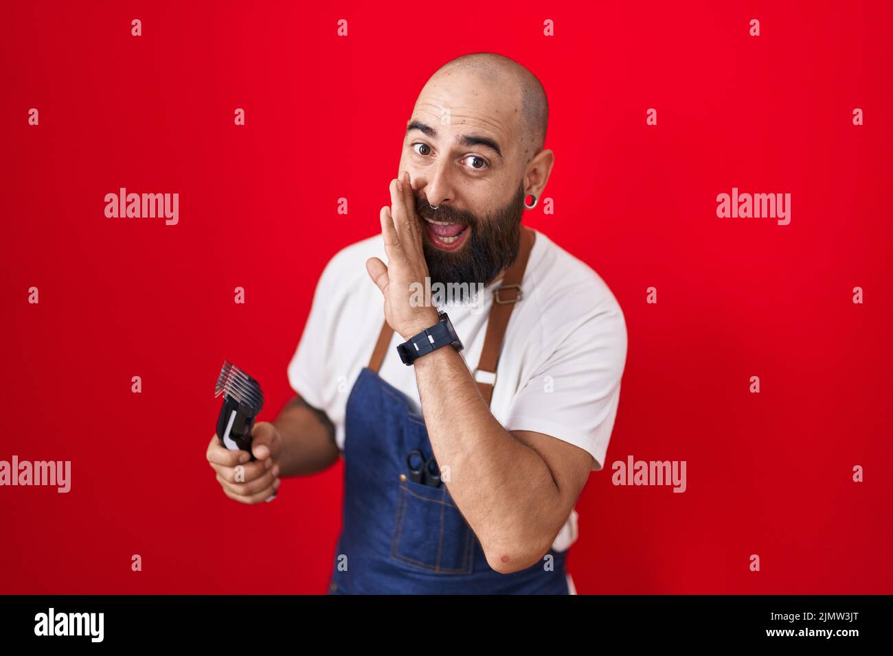 Young hispanic man with beard and tattoos wearing barber apron holding ...