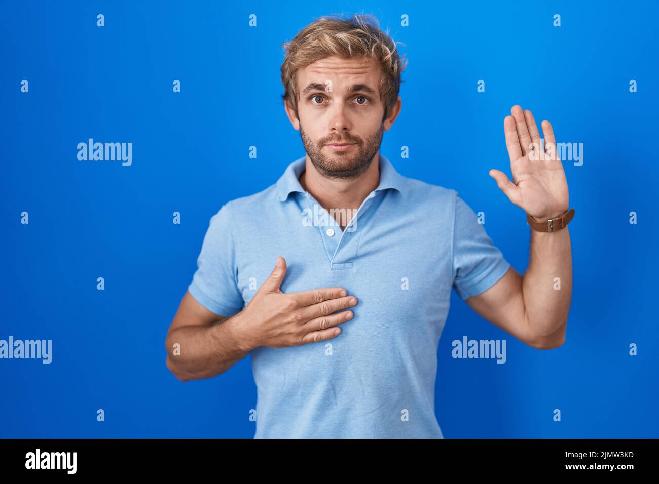 Caucasian man standing over blue background swearing with hand on chest ...