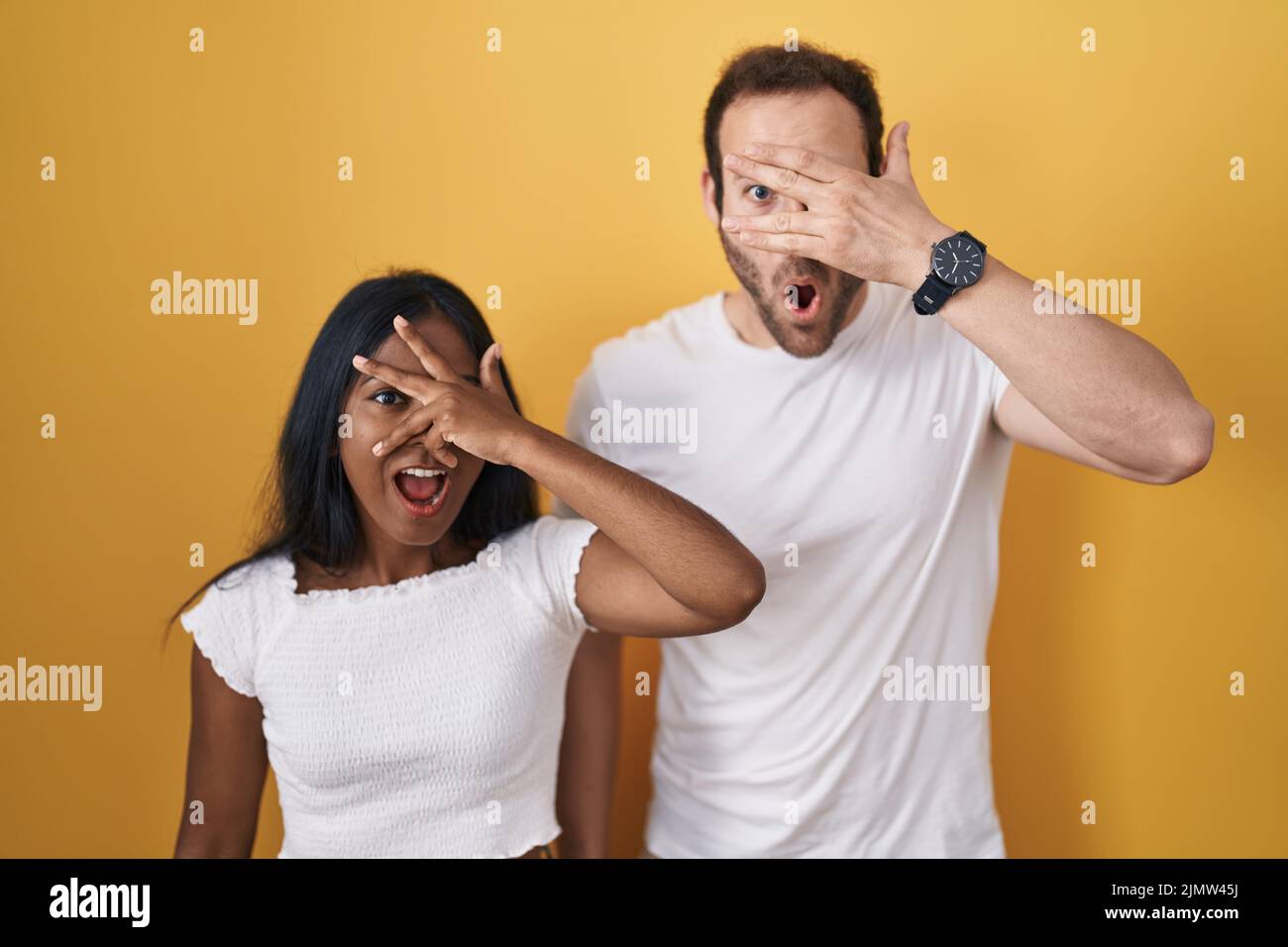 Interracial couple standing over yellow background peeking in shock ...