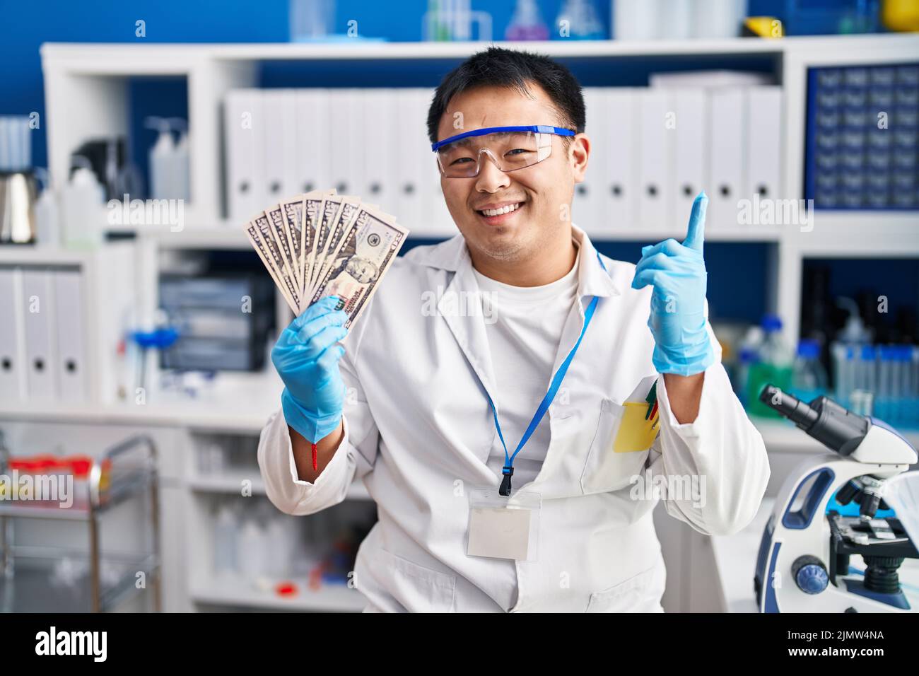Young chinese man working at scientist laboratory holding money smiling happy pointing with hand ...