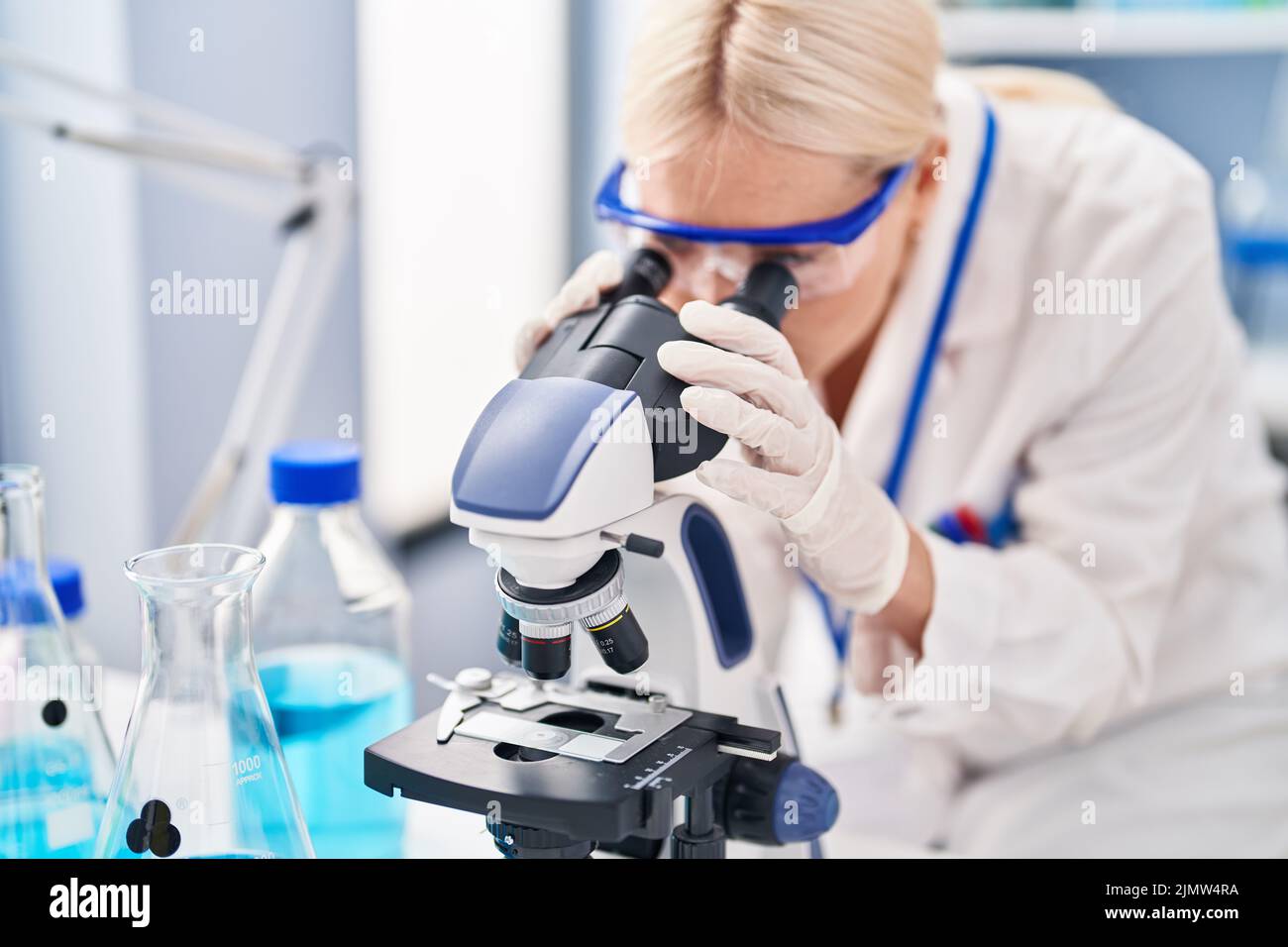 Young blonde woman wearing scientist uniform using microscope at ...