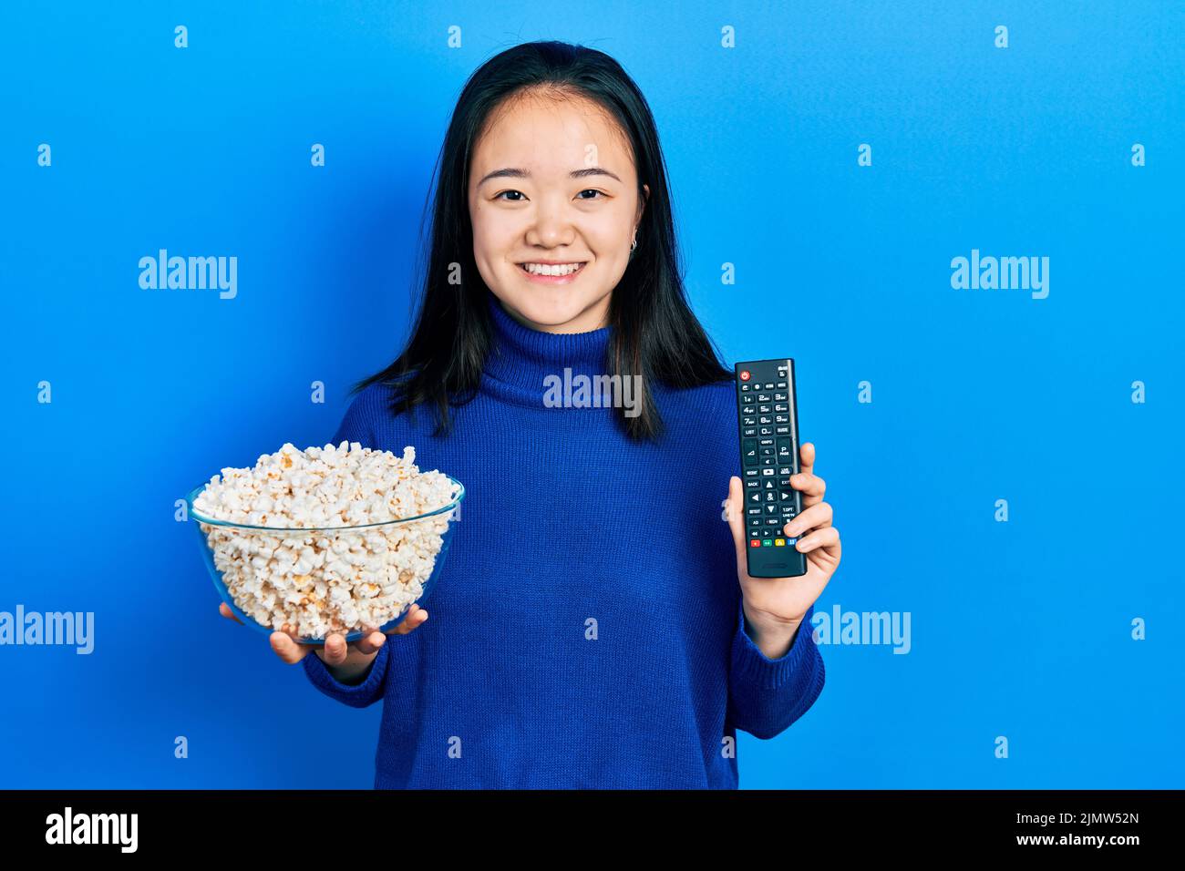 Young chinese girl eating popcorn using tv control smiling with a happy ...