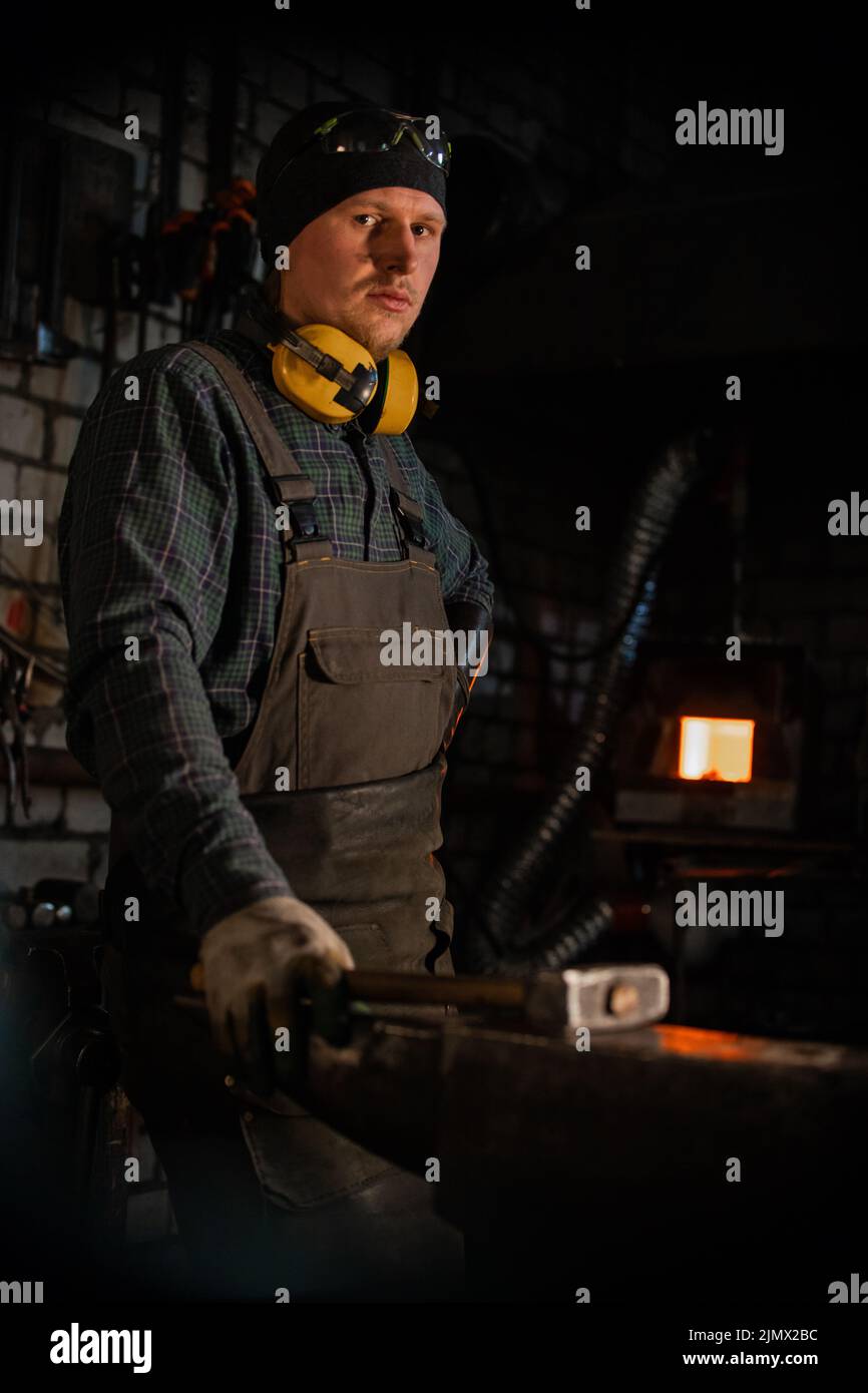 A man blacksmith standing in dark workshop holding a hammer on the ...