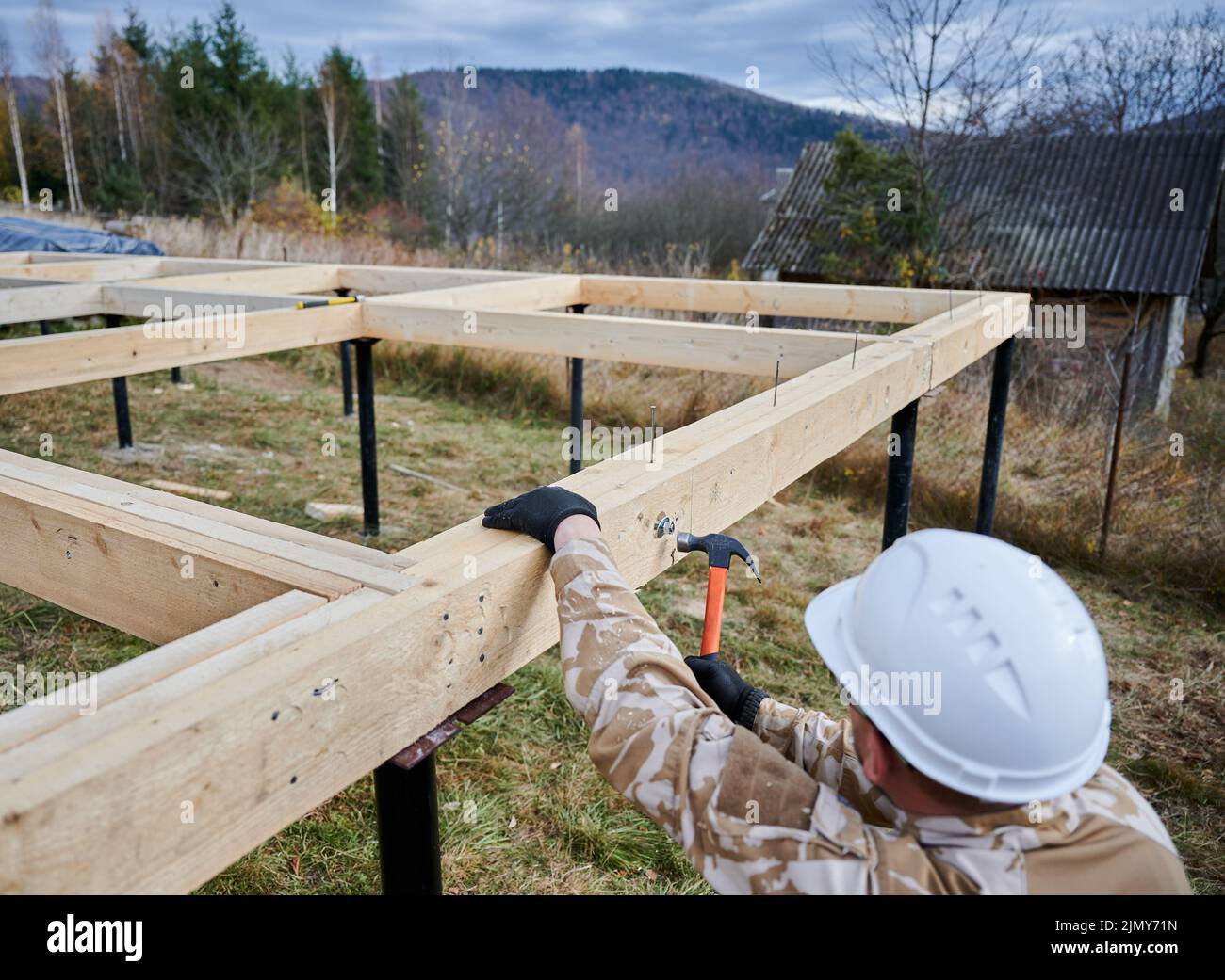 Man worker building wooden frame house on pile foundation. Carpenter hammering bolt into wooden ...