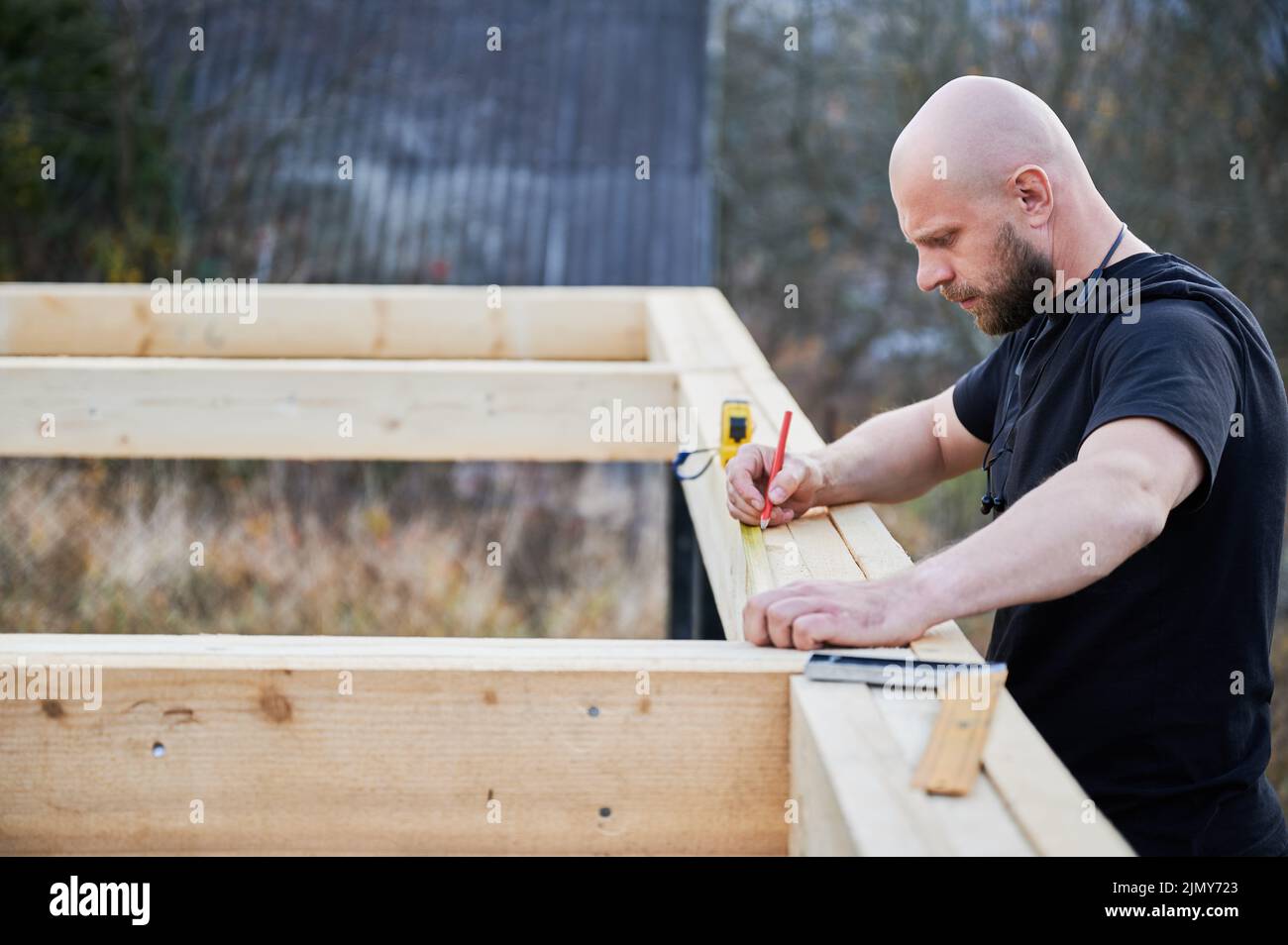 Man worker building wooden frame house on pile foundation. Bald carpenter using tape measure for ...