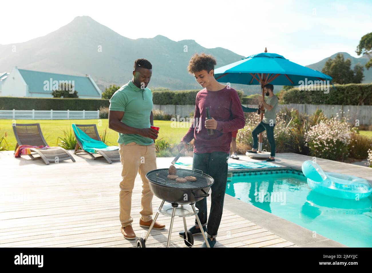 Male multiracial friends having beer and cooking meat on barbecue grill at poolside in summer ...