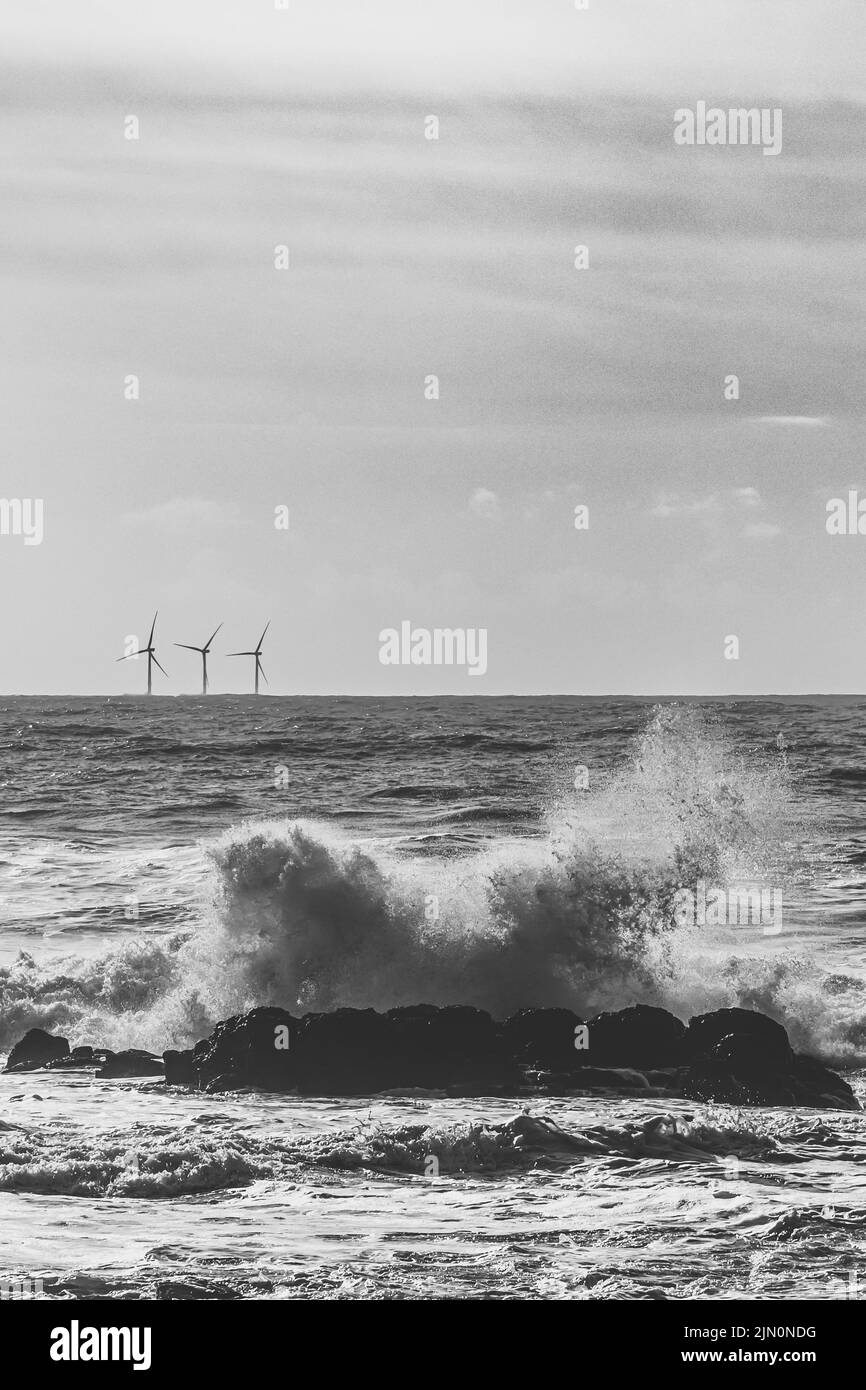 A vertical grayscale shot of the sea waves with wind turbines in the ...