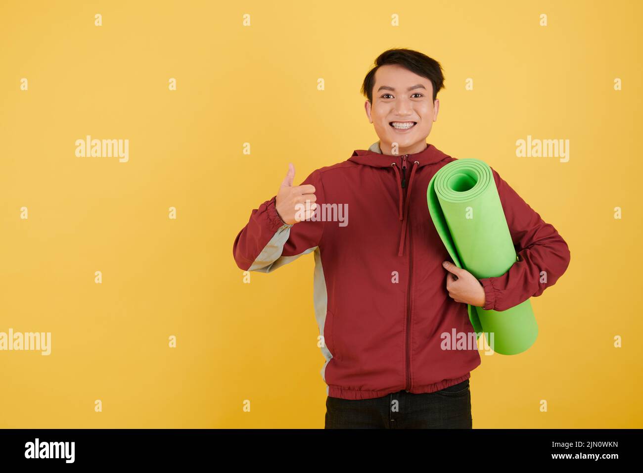 Portrait of excited young man holding yoga mat and showing thumbs-up ...
