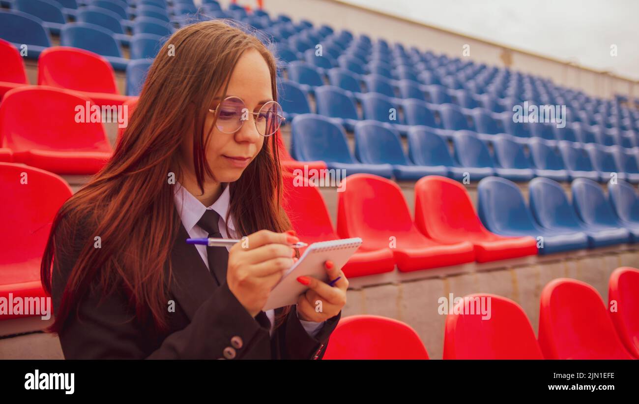 Young woman in glasses with notepad, pen sitting on stadium bleachers ...