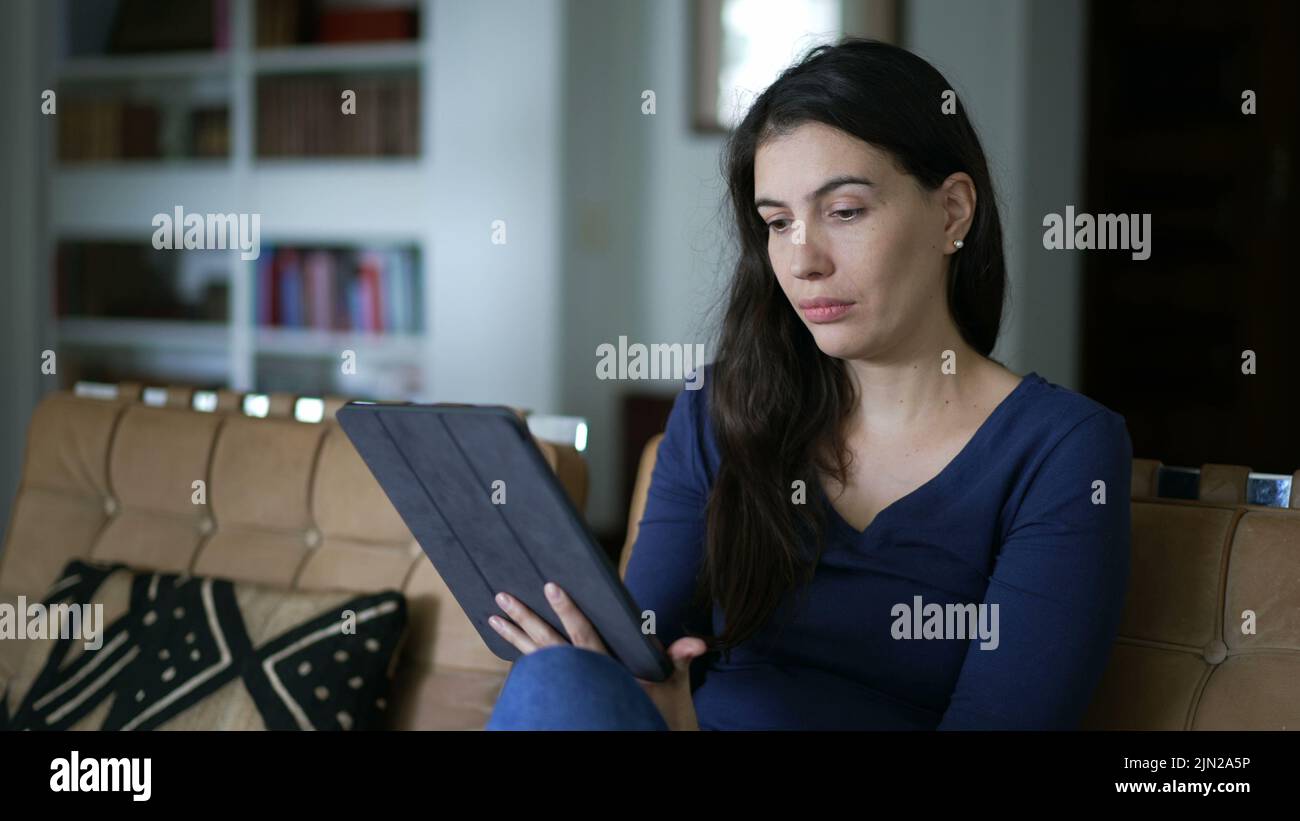 Woman sitting at living room couch staring at tablet device screen with ...