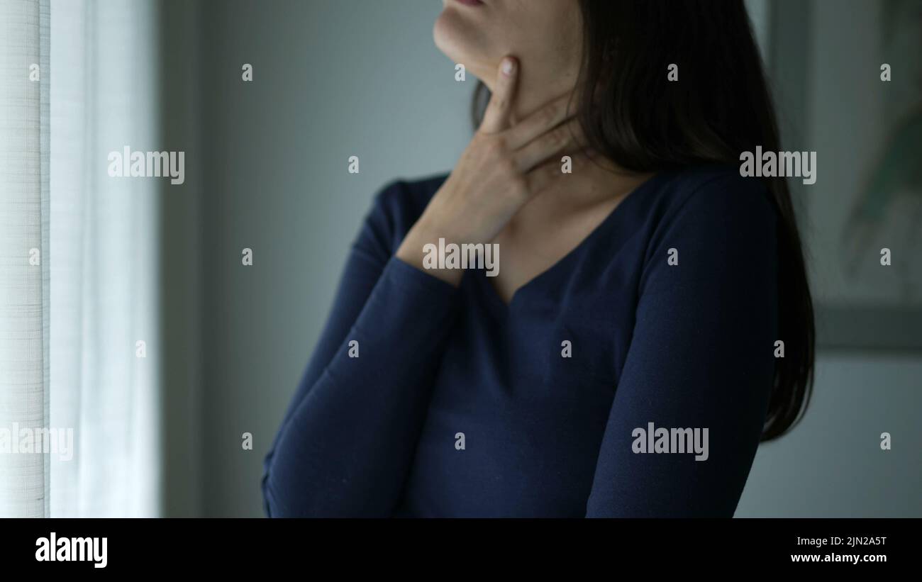 Worried woman standing by window touching her neck and chest seeking comfort. Thoughtful ...
