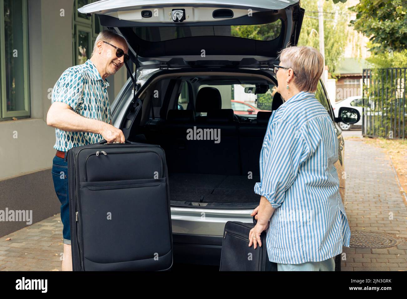 Retired man and woman loading luggage in car trunk, preparing to leave ...
