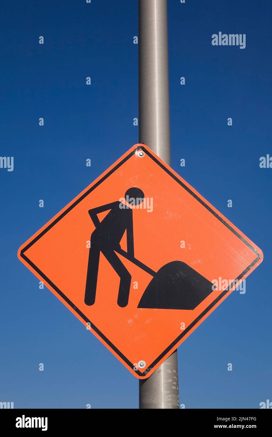 Orange pictogram Men at Work warning sign on metal pole against a blue ...