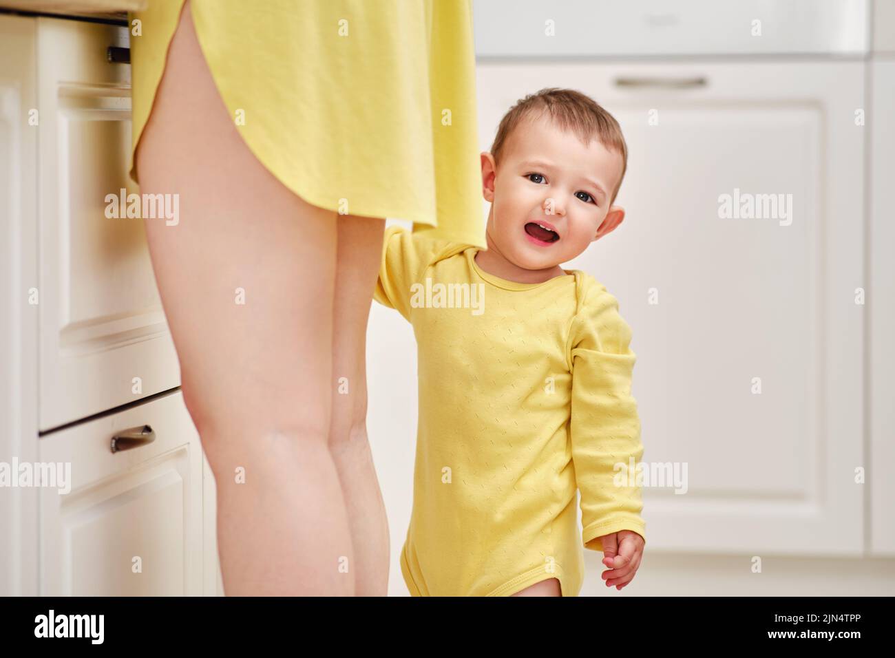 Unhappy toddler baby boy stands holding his mother legs in the kitchen ...