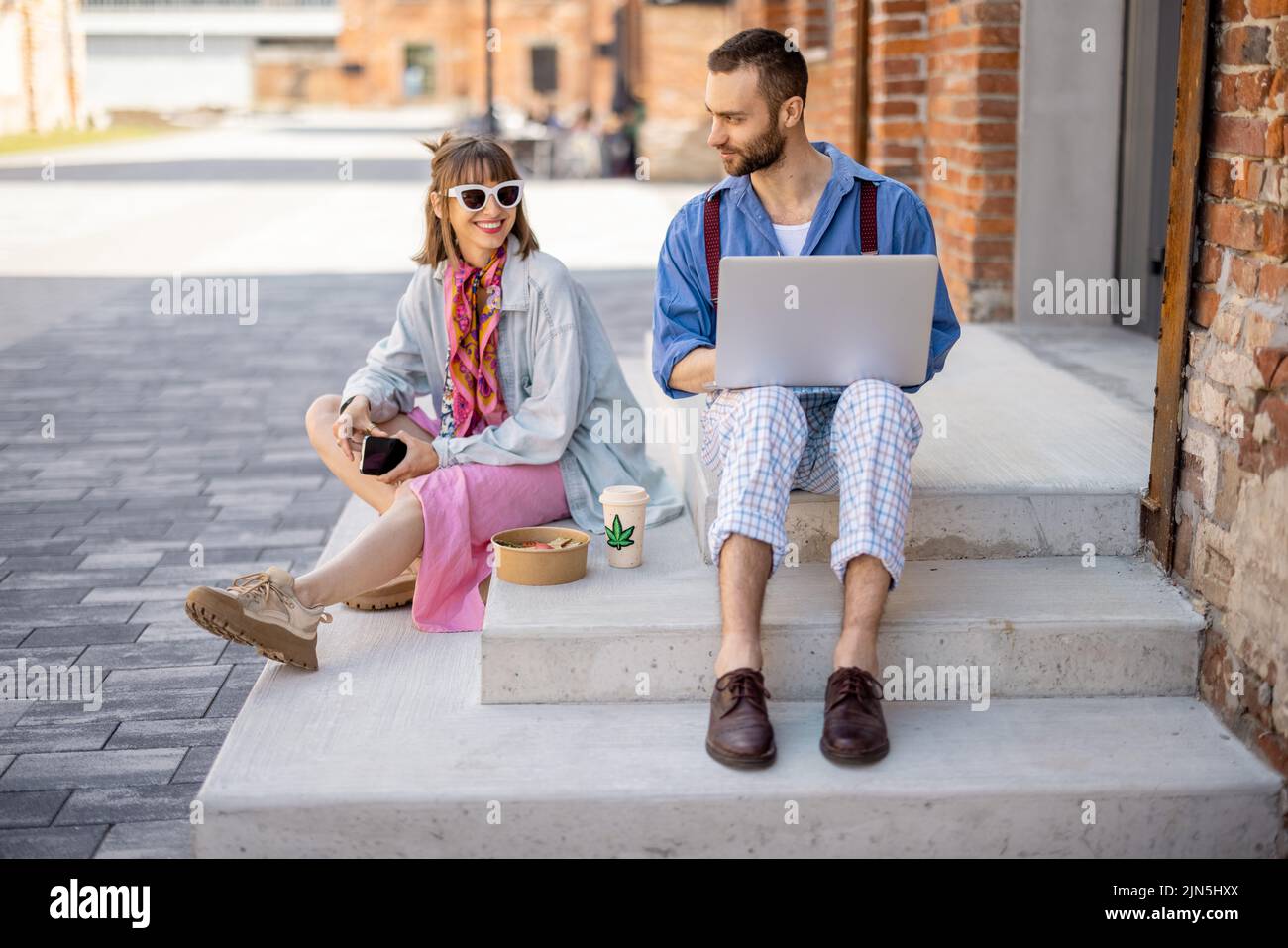 Stylish colleagues work on laptop and eat take away food on a street ...