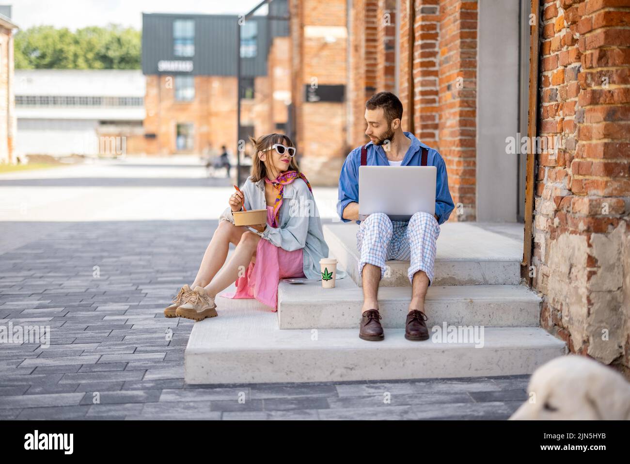 Stylish colleagues work on laptop and eat take away food on a street ...